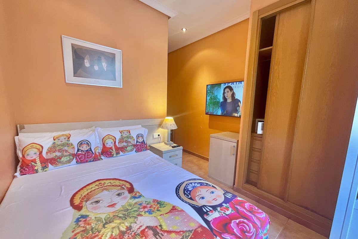 Quarto de hotel com uma cama adornada com almofadas de bonecas russas Matrioshka, uma televisão na parede e um guarda-roupa de madeira.