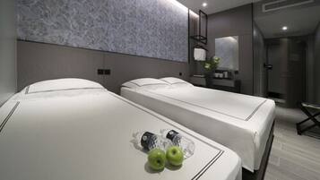 Quarto de hotel com duas camas, parede com padrão marmorizado, uma pequena mesa com um vaso e duas maçãs, e um espelho sobre a mesa.