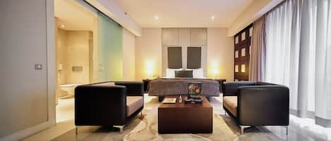 Quarto de hotel moderno com uma cama grande, duas poltronas, uma mesa de centro e vista para a cidade pela janela.