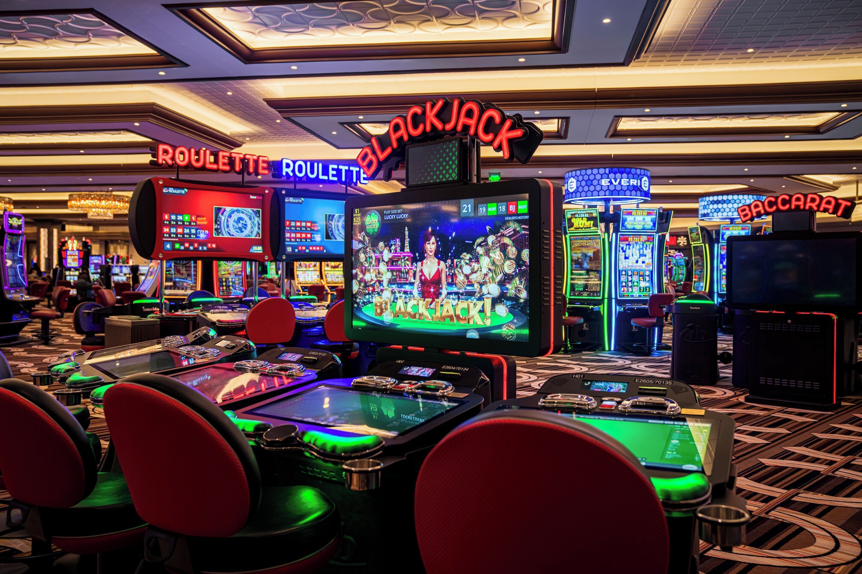 Um cassino com mesas de roleta e blackjack, uma tela grande exibindo um jogo, e um aviso para baccarat.
