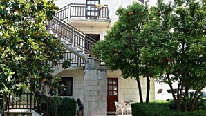 Um edifício de dois andares com varanda, escadaria e um jardim com árvores e uma mesa.