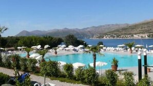 Uma área à beira da piscina com espreguiçadeiras e guarda-sóis, vista desimpedida para o mar, e montanhas ao fundo.