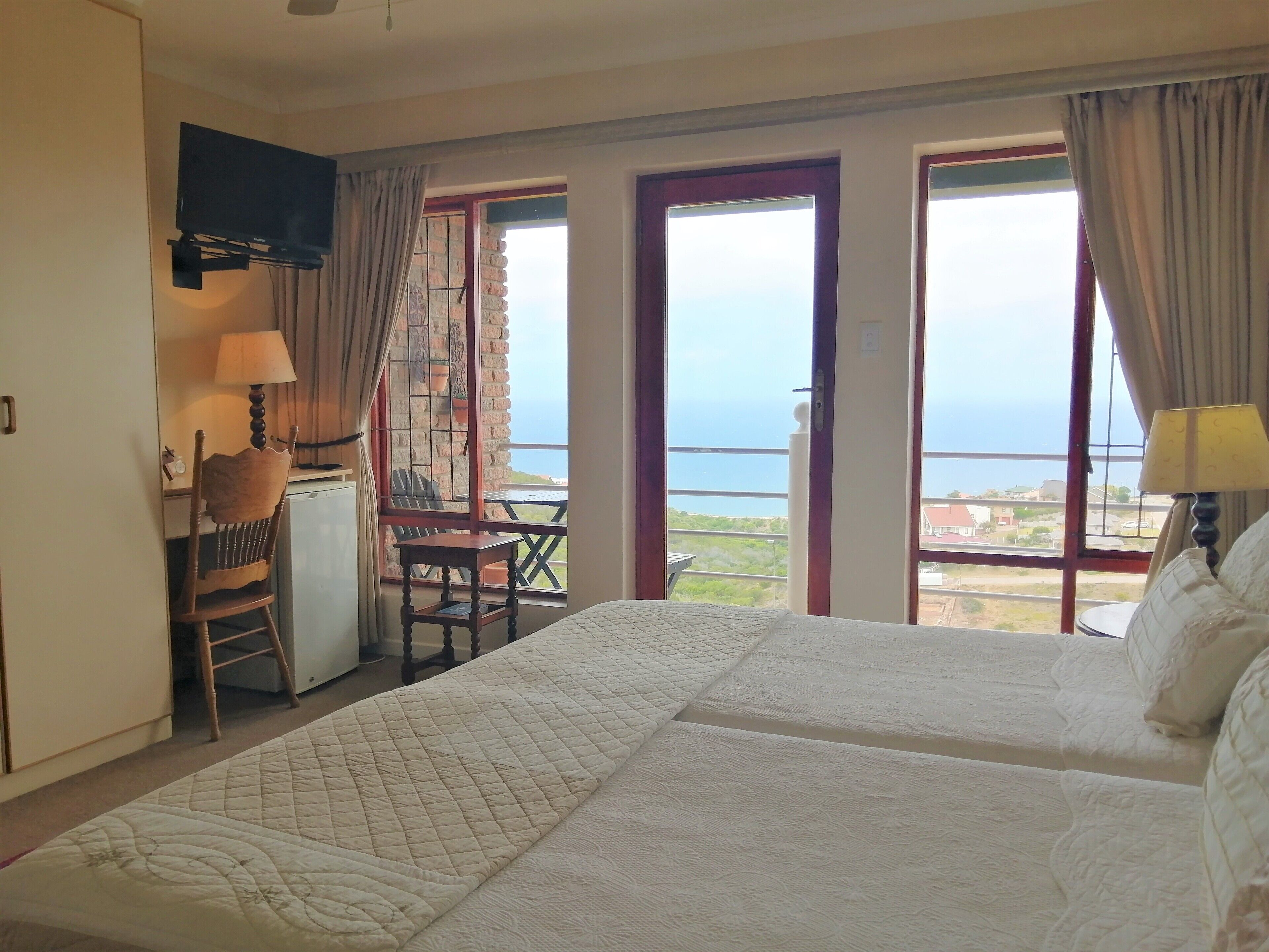 Quarto de hotel com uma cama, uma escrivaninha, uma cadeira e uma mesa. Há duas janelas com cortinas, e é visível uma vista para o mar.