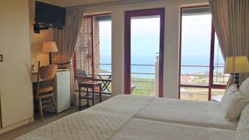 Quarto de hotel com uma cama, uma escrivaninha, uma cadeira e uma mesa. Há duas janelas com cortinas, e é visível uma vista para o mar.
