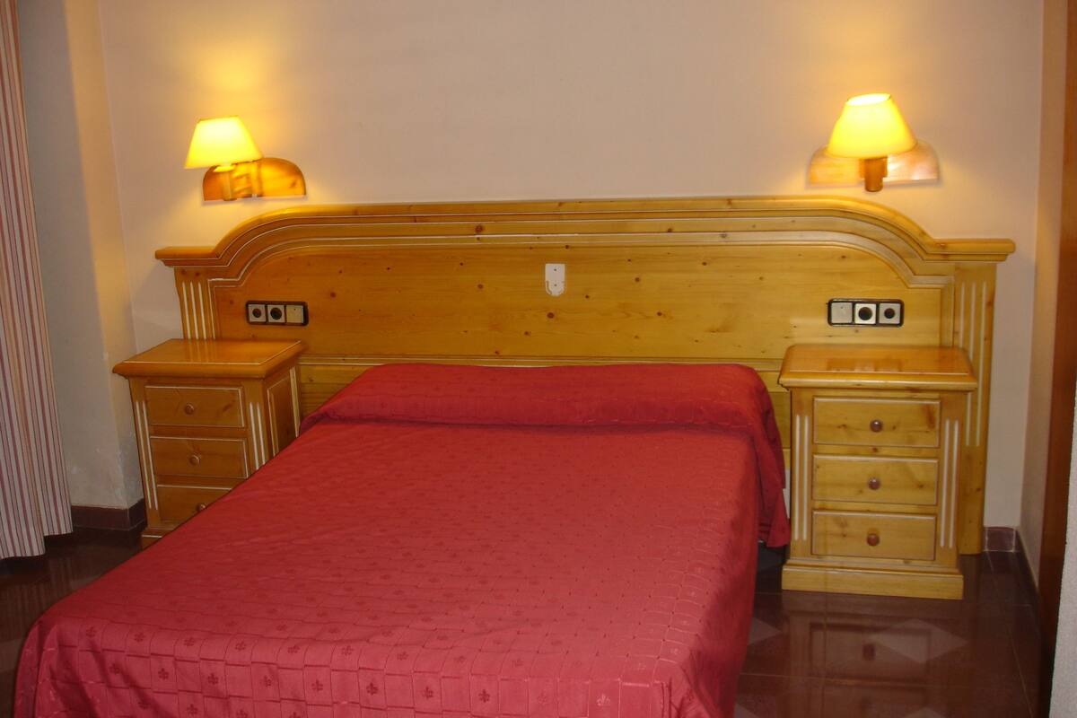 Quarto de hotel padrão com cabeceira de madeira, duas mesas de cabeceira com abajures, colcha vermelha e uma parede lisa.
