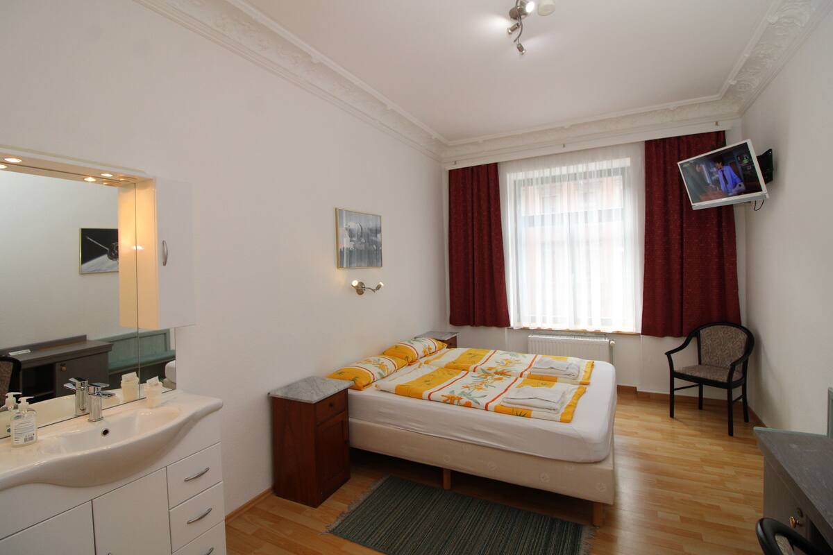 Quarto com uma cama, mesa de cabeceira, espelho, televisão na parede e janela com cortinas.