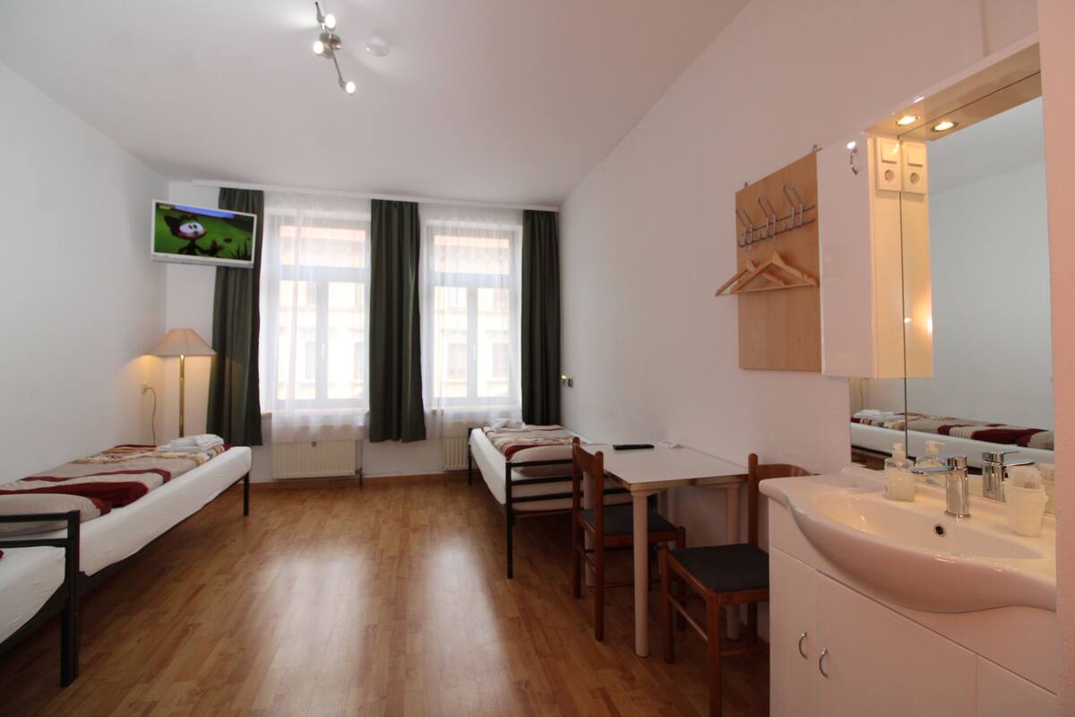 Quarto com cama, escrivaninha, cadeira, mesa, televisão, espelho e pia.