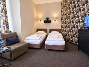 Quarto de hotel com duas camas, um sofá, uma mesinha com uma chávena e garrafas de água, e uma televisão num móvel.