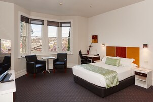 Quarto de hotel com cama, escrivaninha, cadeira e vista da cidade através de grandes janelas.