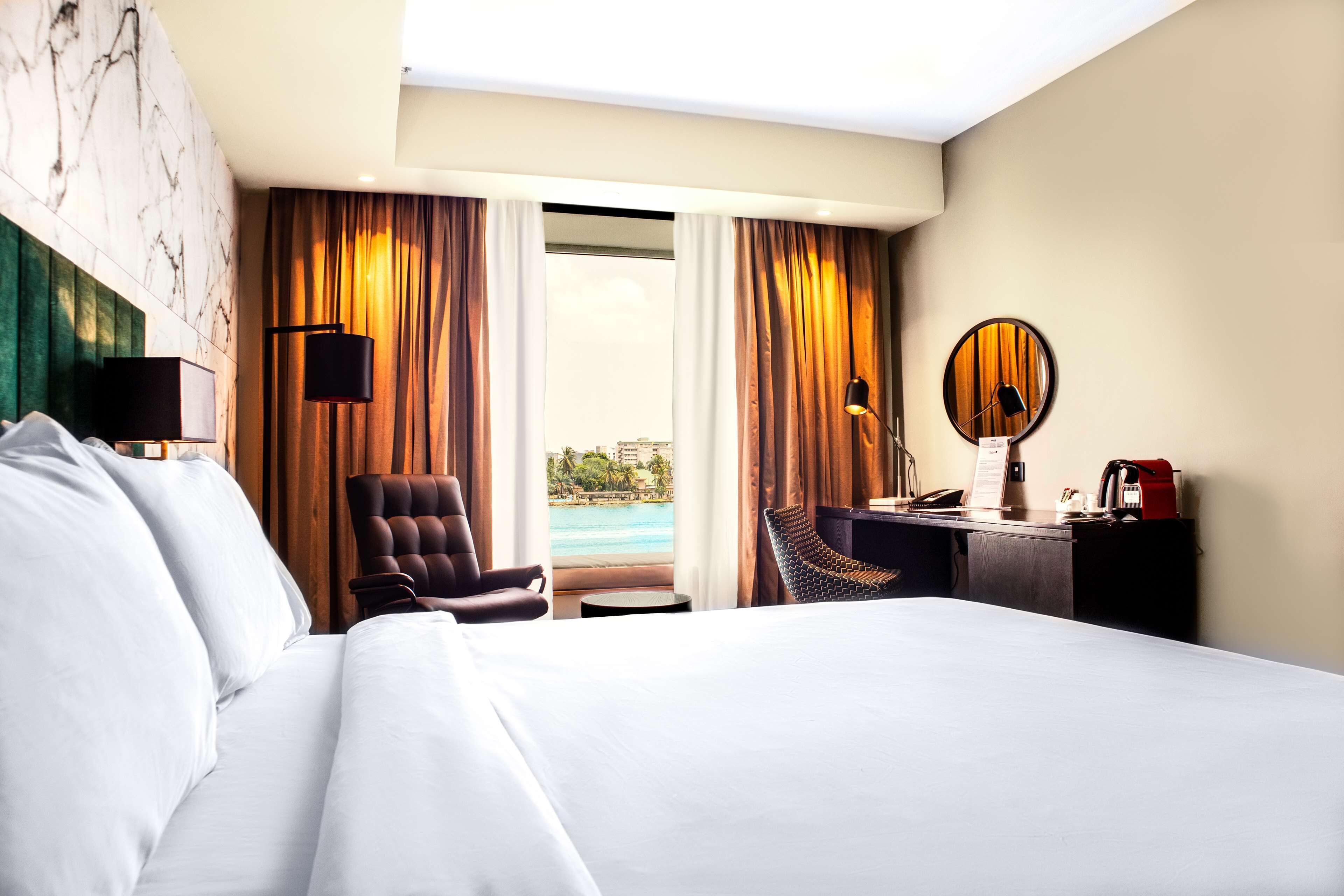 Quarto de hotel com uma cama grande, uma escrivaninha com cadeira, uma mesinha, uma luminária, um espelho e vista para a praia e oceano pela janela.