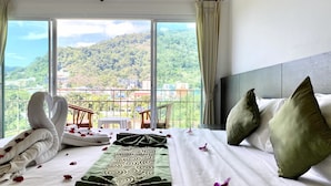 Quarto de hotel com cama, almofadas, mesa e varanda com vista para uma colina arborizada.