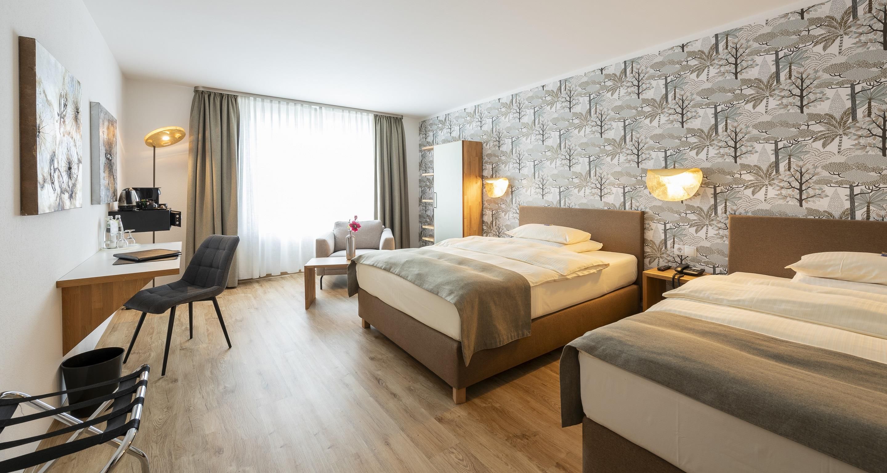 Quarto de hotel com duas camas, uma escrivaninha, uma cadeira e uma janela grande com cortinas.