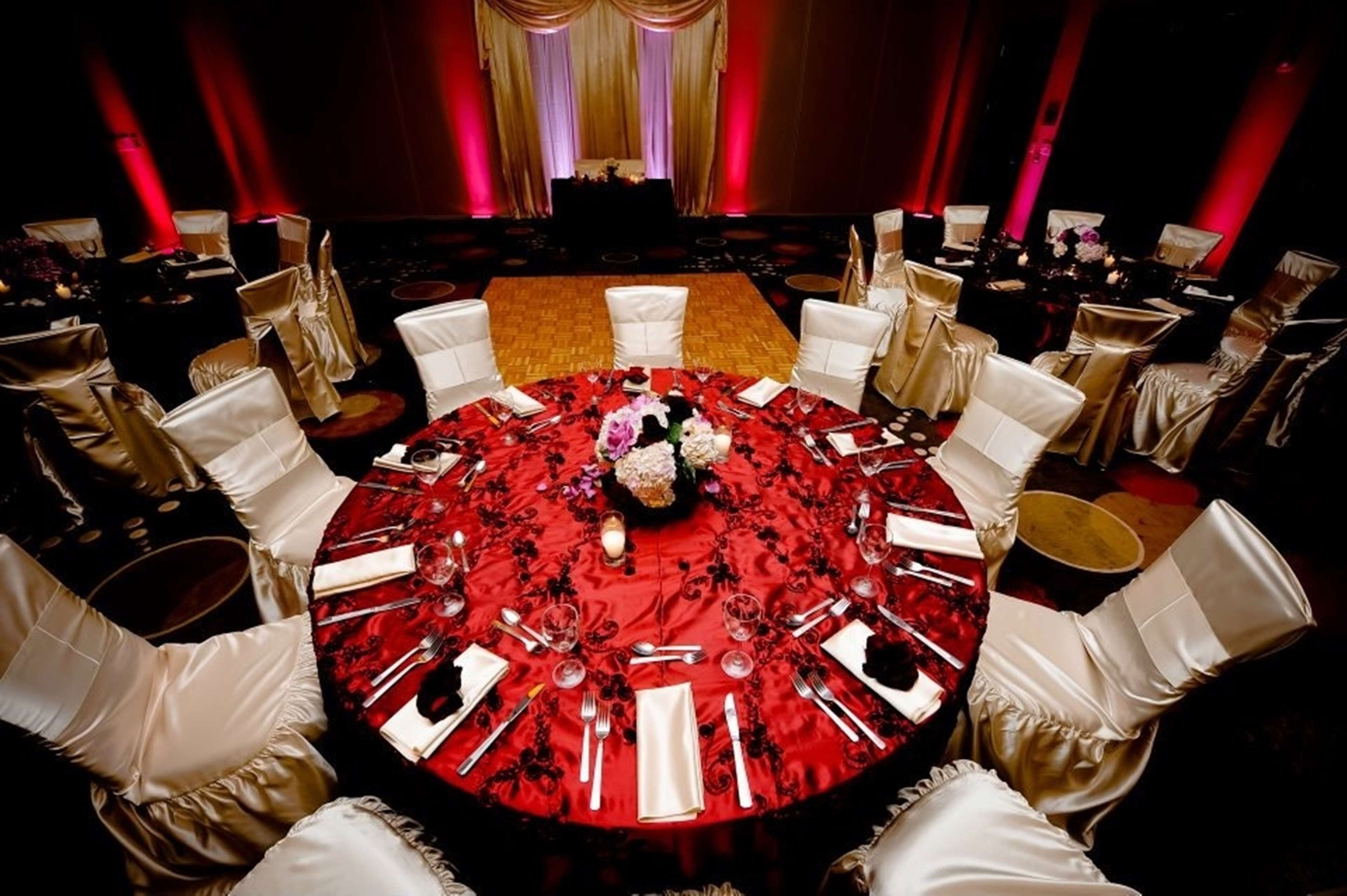 Mesa redonda com toalha vermelha, rodeada por cadeiras brancas com fitas douradas, preparada para um evento formal.