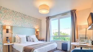 Quarto de hotel com uma cama grande, mesas de cabeceira com abajures, uma mesinha com uma cadeira, uma janela com cortinas e uma televisão na parede.