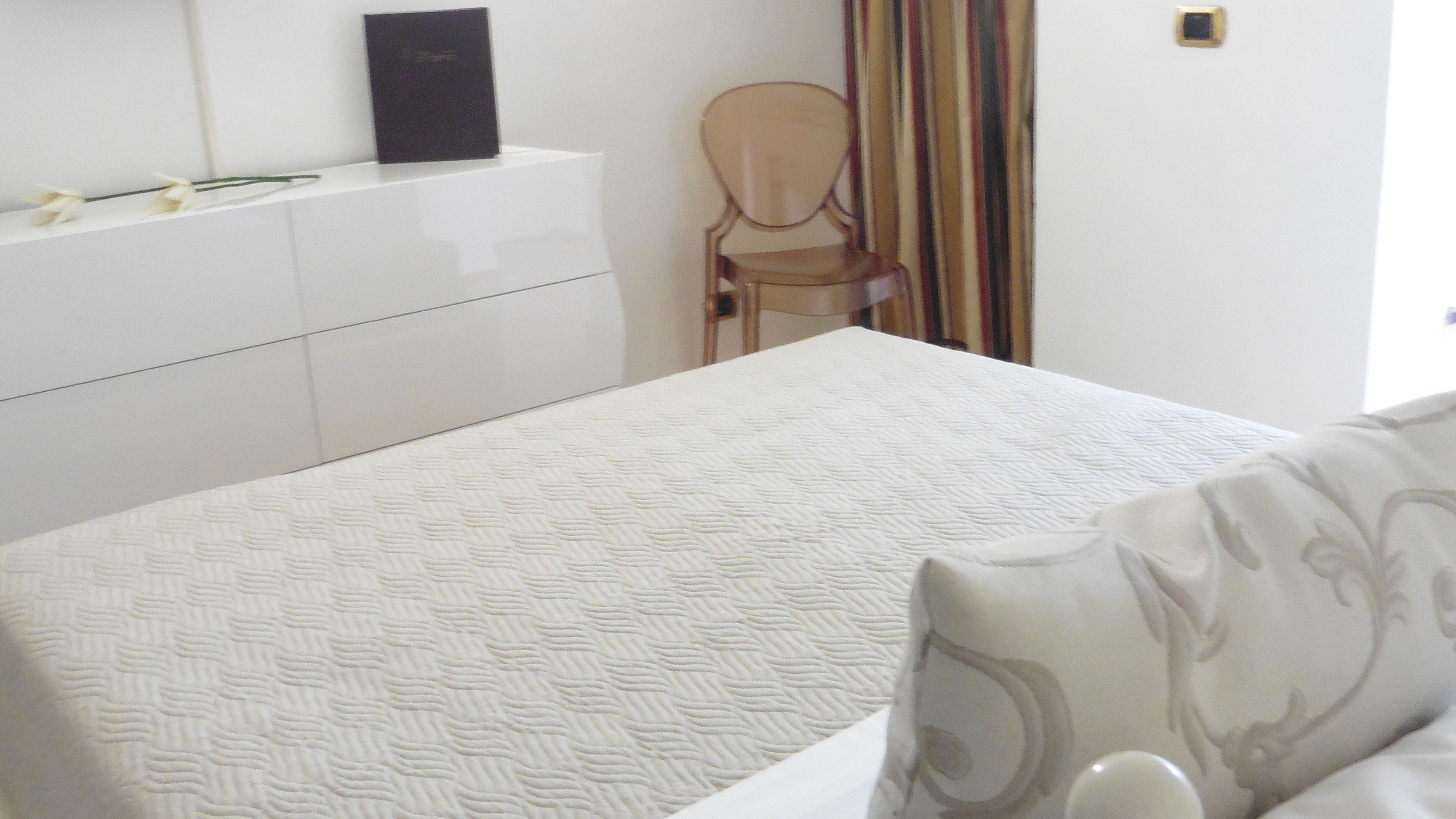 Quarto de hotel com uma cama, uma cadeira, cômoda branca e cortina.