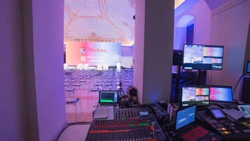 Sala de controle com vários monitores, equipamentos de áudio e uma tela grande exibindo 'TOTAL The European House Ambrosetti'.