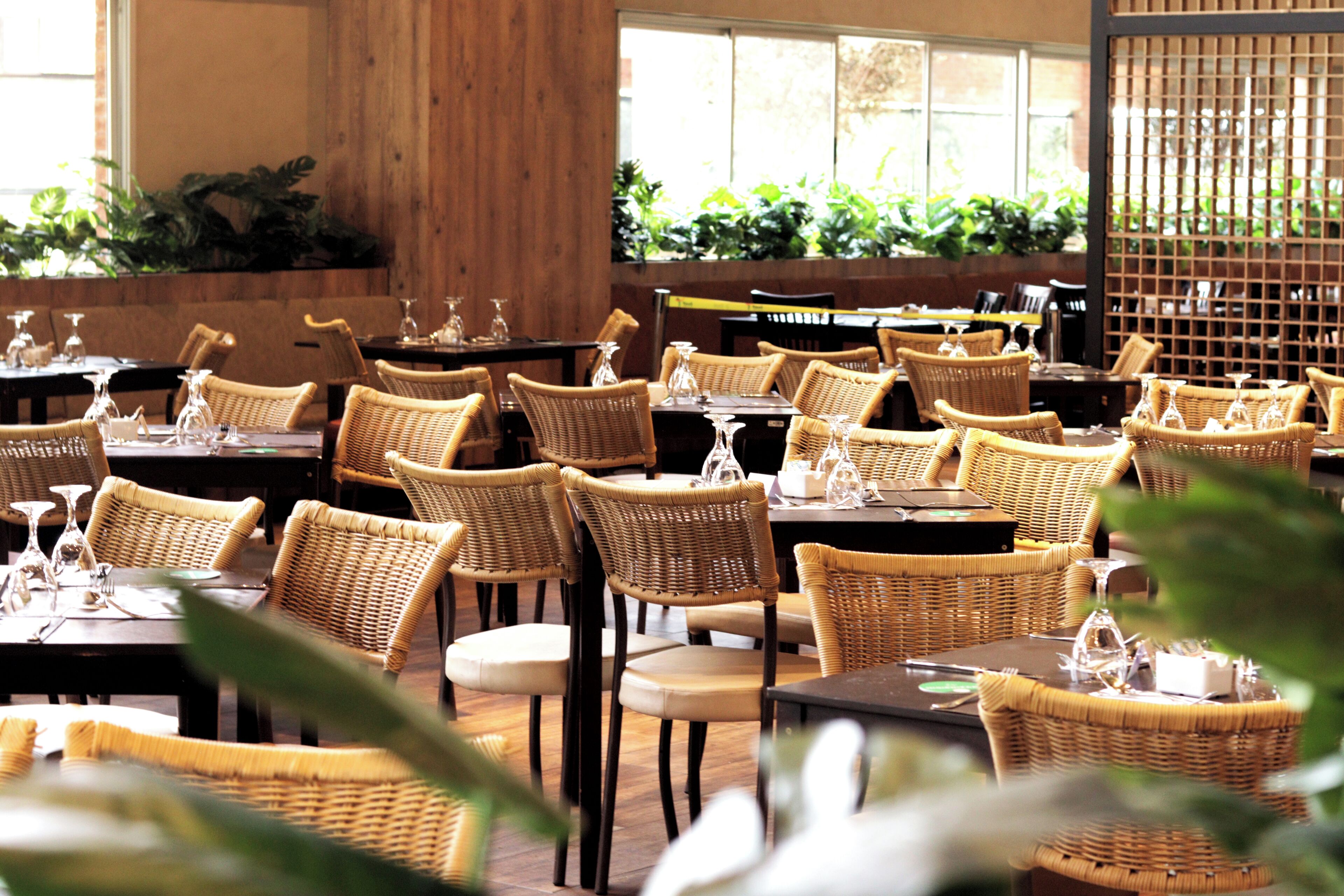 Um restaurante com mobiliário de vime, mesas preparadas com copos e vista para o verde lá fora.