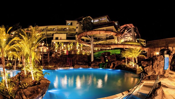 Piscina noturna com toboá, rodeada por plantas tropicais e um edifício com vários andares.