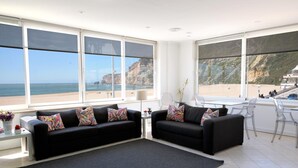 Uma sala de estar moderna com dois sofás pretos, um tapete cinzento e grandes janelas com vista para uma praia.