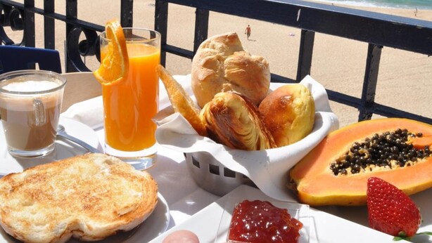 Mesa de café da manhã com croissants, um copo de suco de laranja, uma fatia de torrada, um prato de presunto, queijo e uma salada de frutas em uma varanda com vista para a praia.