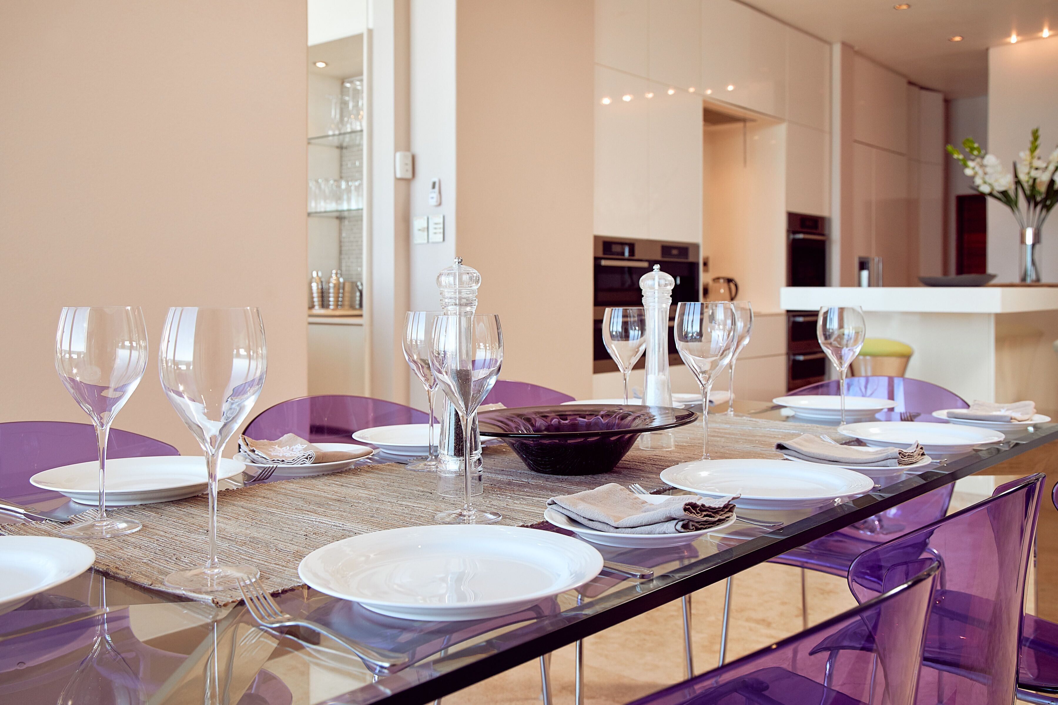 Mesa de jantar posta com pratos brancos, copos de vinho transparentes e cadeiras roxas em uma cozinha moderna com cores neutras.