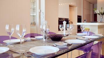 Mesa de jantar posta com pratos brancos, copos de vinho transparentes e cadeiras roxas em uma cozinha moderna com cores neutras.