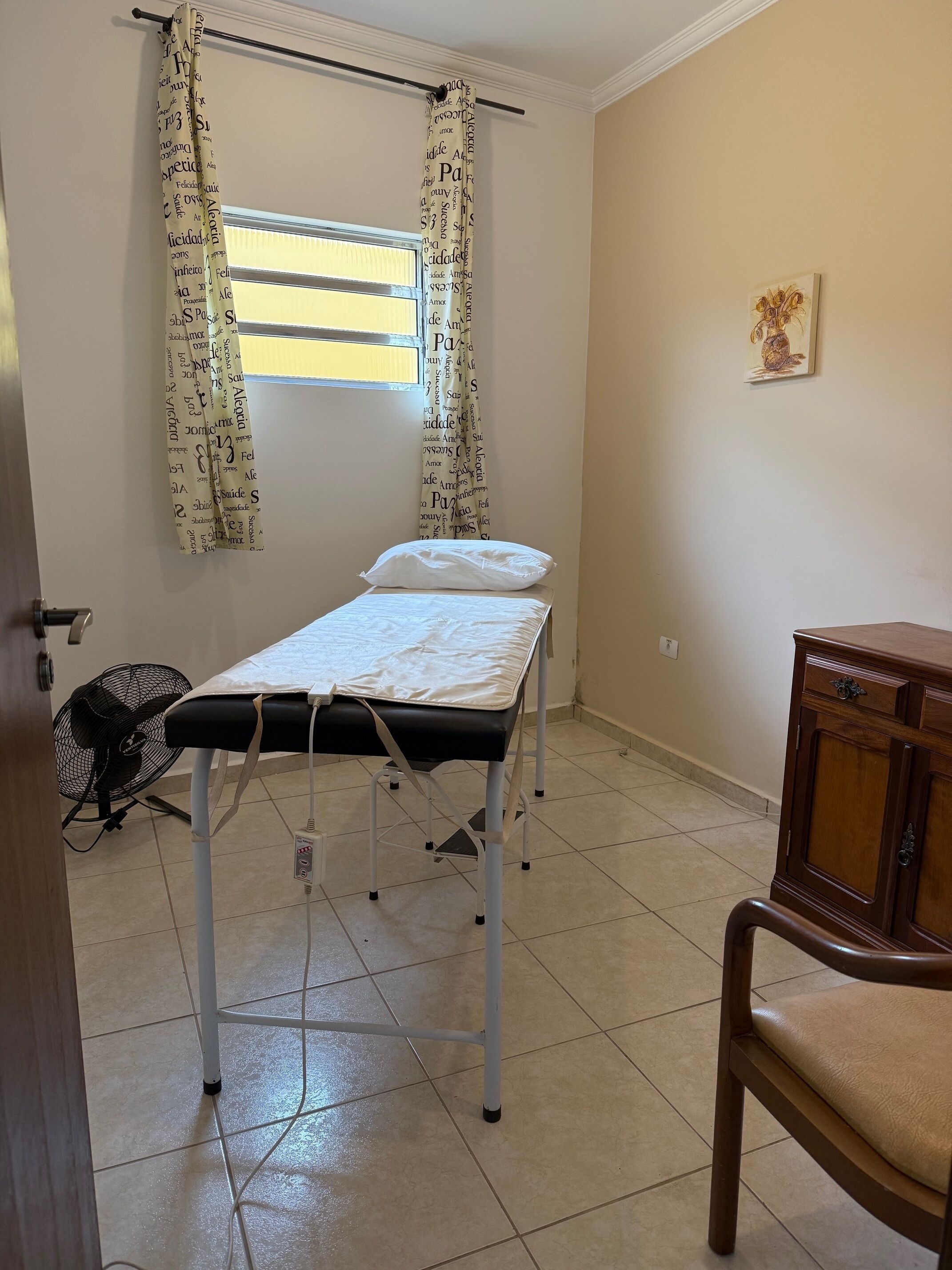 Quarto com cama, cadeira, ventoinha e janela com cortinas.