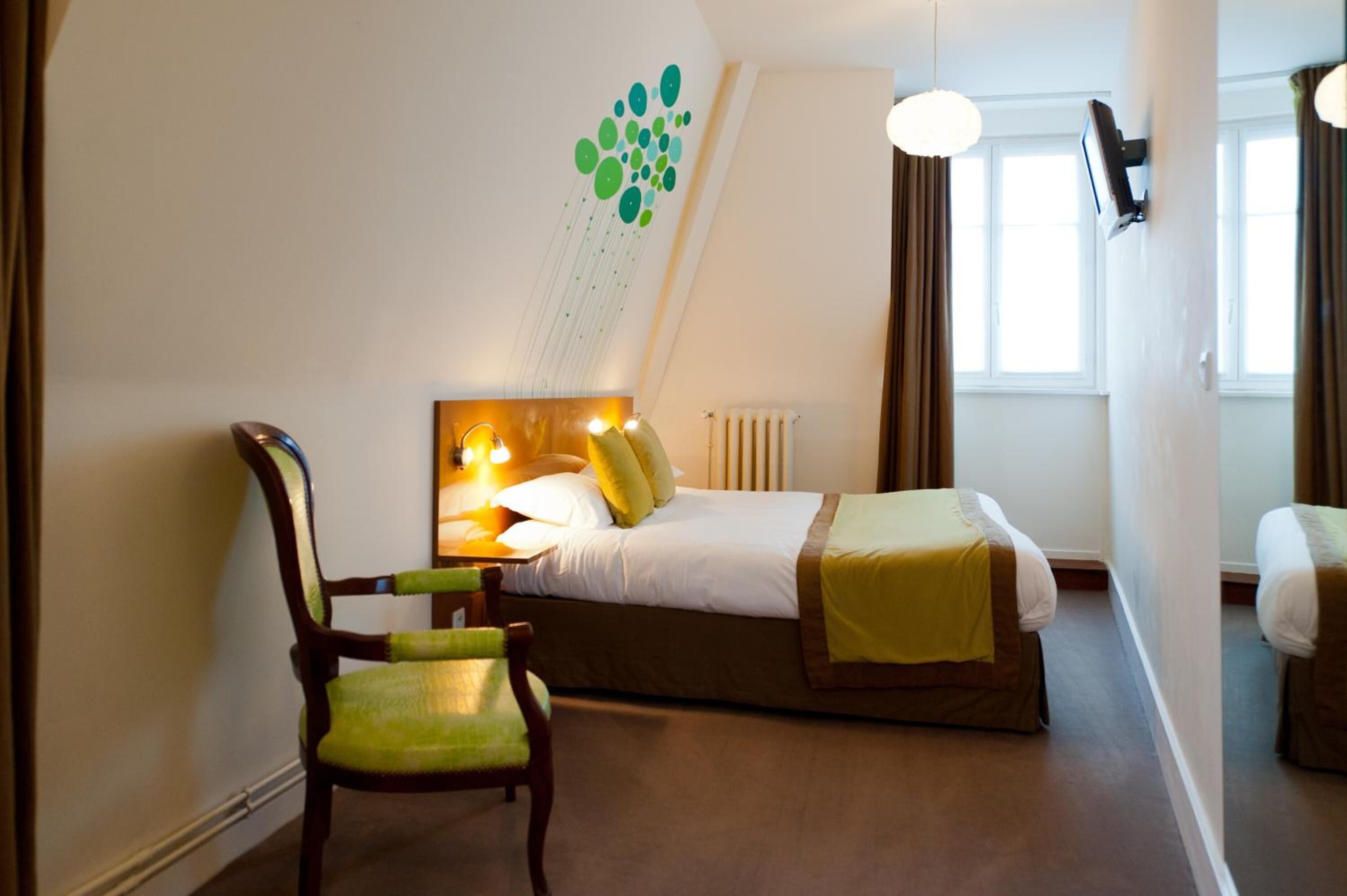 Quarto de hotel com cama, uma cadeira, TV fixada na parede e janela com cortinas.