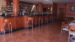 Uma área de bar com balcões e banquetas de madeira, um espaço de estar com mesas e cadeiras, e uma parede de tijolos ao fundo.