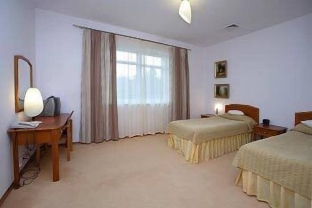 Quarto de hotel com duas camas, uma escrivaninha, uma cadeira, um abajur e janela com cortinas.