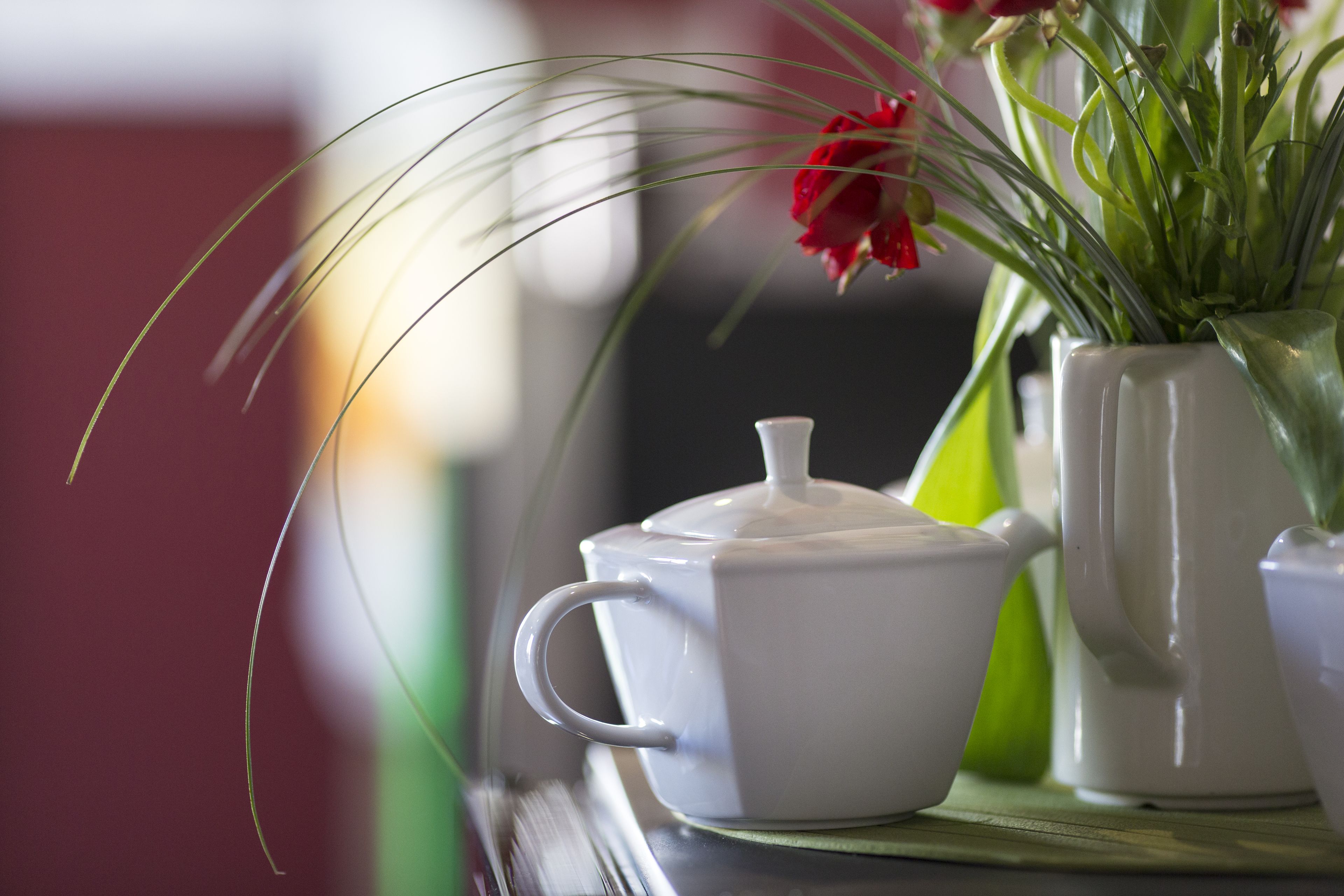 Uma chaleira branca e um açucareiro branco sobre uma mesa com um vaso verde contendo flores vermelhas.