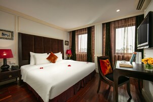 Quarto de hotel com uma cama grande, uma escrivaninha com cadeira, uma televisão e uma janela com cortinas.