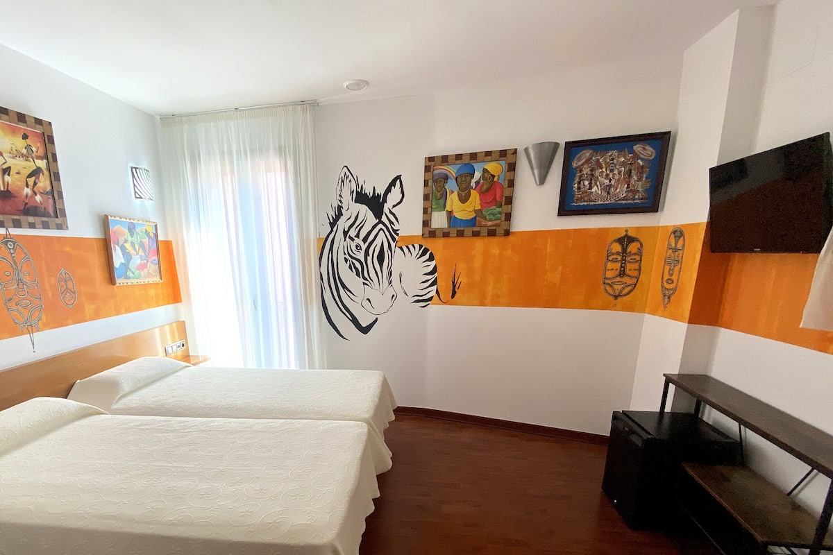 Quarto de hotel com duas camas, um grande mural de zebra e diversas pinturas culturais nas paredes.