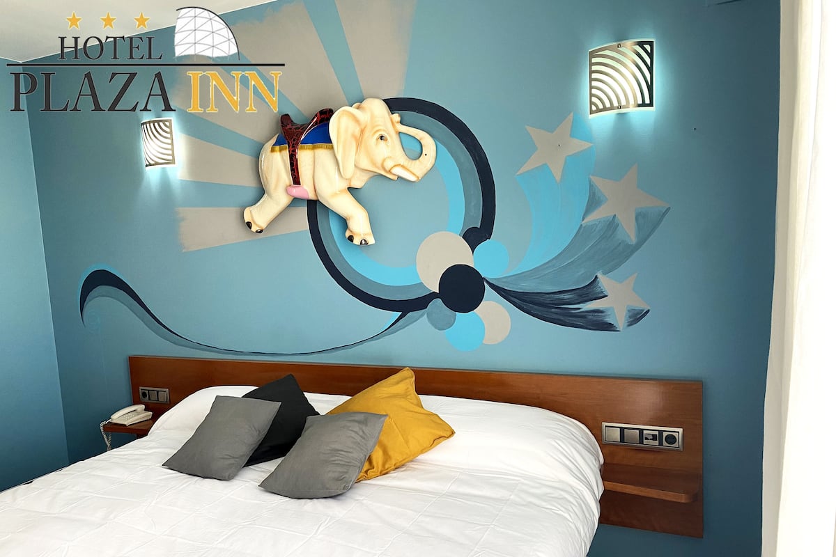 Quarto de hotel com cama, almofadas, mesa de cabeceira, telefone e um mural na parede.