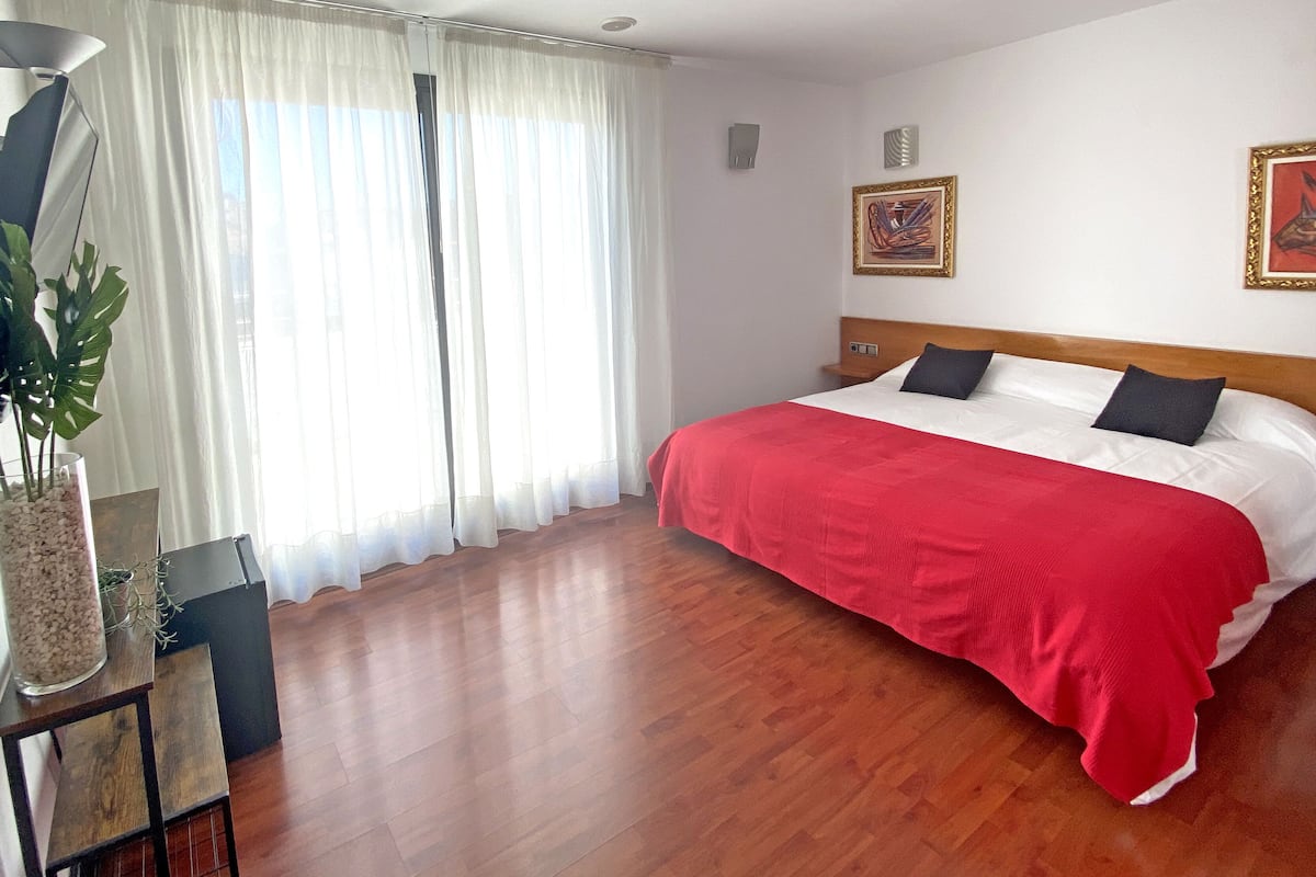 Quarto de hotel com piso de madeira, uma cama grande com manta vermelha, mesa de cabeceira, luminária e um vaso de planta.