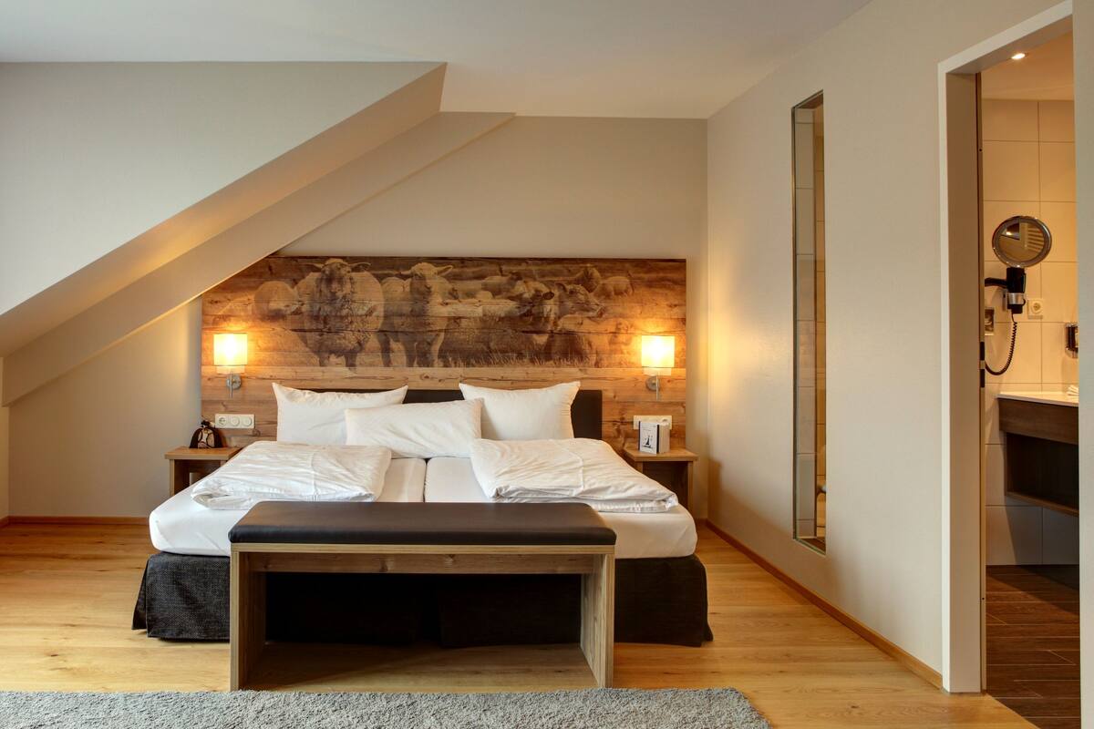 Um quarto com teto inclinado, uma cama grande com roupa de cama branca, duas mesas de cabeceira com abajures e uma cabeceira de madeira com um mural.