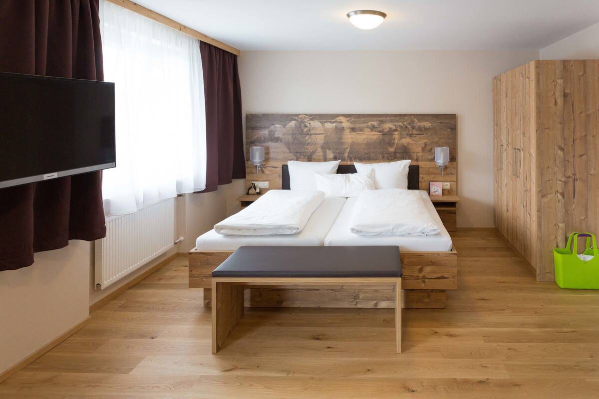 Quarto de hotel com uma cama, um banco, uma TV e cabeceira de madeira.
