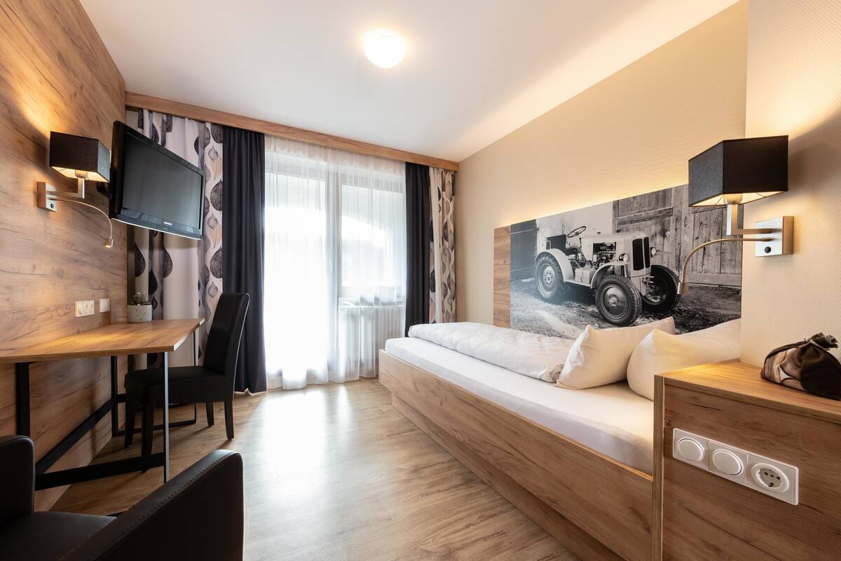 Quarto de hotel moderno com escrivaninha de madeira, cama com cabeceira, televisão fixada na parede e janela grande com cortinas.