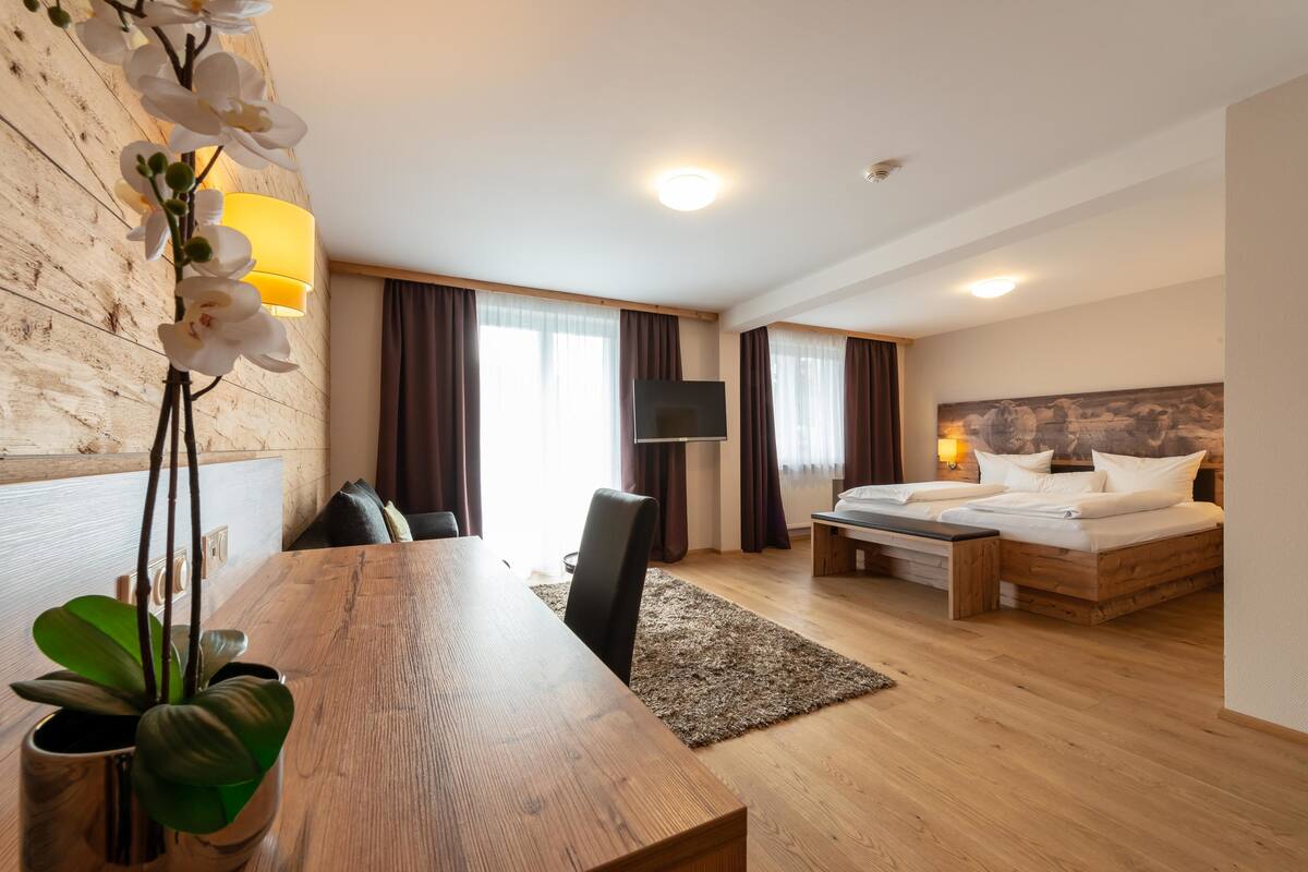 Quarto de hotel moderno com uma mesa de jantar de madeira, cama com encosto, televisão de tela plana e uma janela grande com cortinas.
