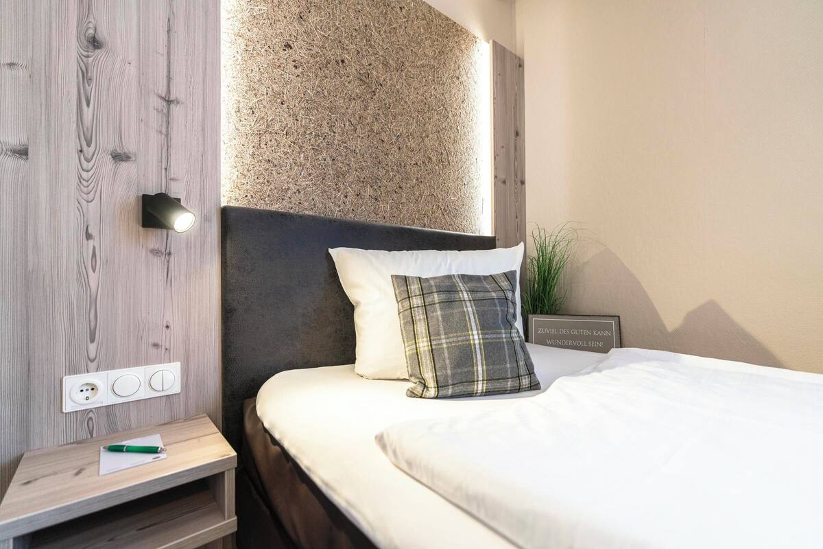 Cama arrumada com travesseiro branco e travesseiro xadrez, mesa de cabeceira com caneta e papel, e luminária fixada na parede.