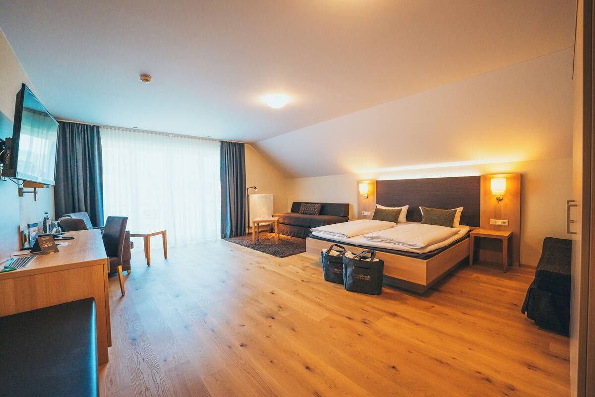 Quarto de hotel com piso de madeira, uma cama com travesseiros, uma escrivaninha com cadeira, televisão e luminária.