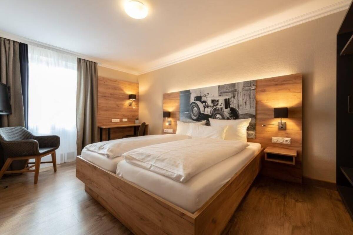 Cama de casal com cabeceira de madeira, mesas de cabeceira com luminárias, uma cadeira e uma janela grande com cortinas.