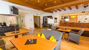 Um hall de hotel moderno com piso de madeira, sofás cinzas, mesas de madeira e uma área de cozinha ao fundo.