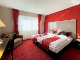 Quarto de hotel com carpete vermelho, duas camas, uma escrivaninha e uma cadeira.