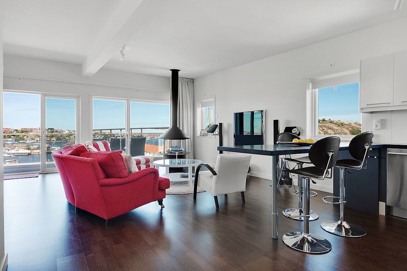 Uma sala de estar moderna com um sofá vermelho, uma poltrona branca e uma área de jantar com banquetas. Grandes janelas proporcionam vista para o mar e edifícios.