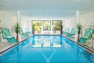 Piscina interna com água azul cristalina, rodeada por espreguiçadeiras e plantas em vasos, com grandes janelas que oferecem vista para o verde.