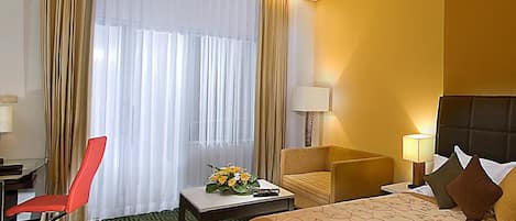 Quarto de hotel com uma cama, uma escrivaninha, uma cadeira, um abajur e uma janela com cortinas.