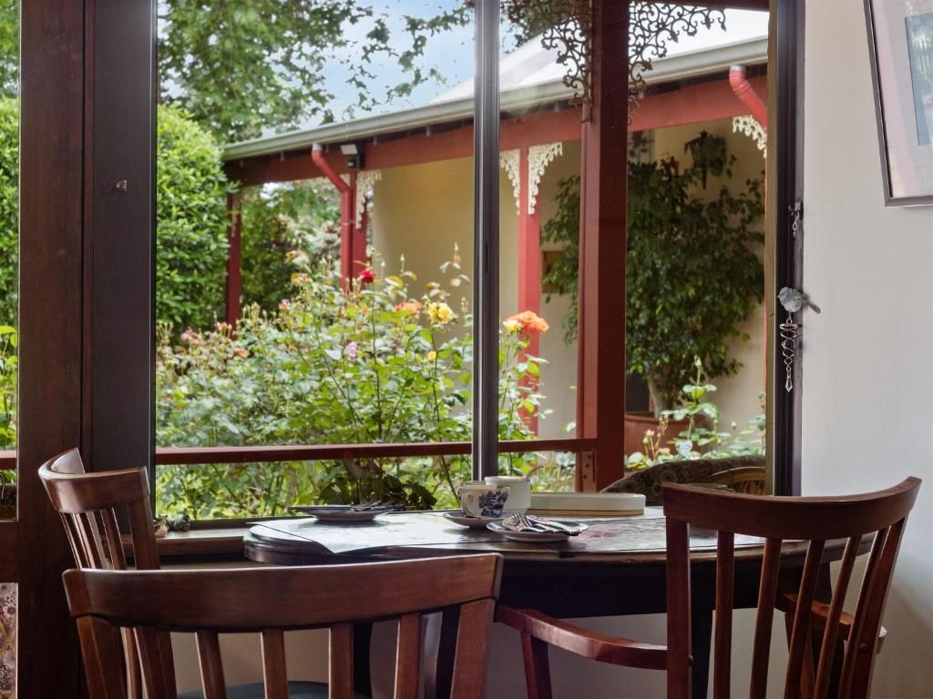 Uma área de refeições com uma mesa posta para dois, uma janela com vista para um jardim e uma estrutura de treliça vermelha.