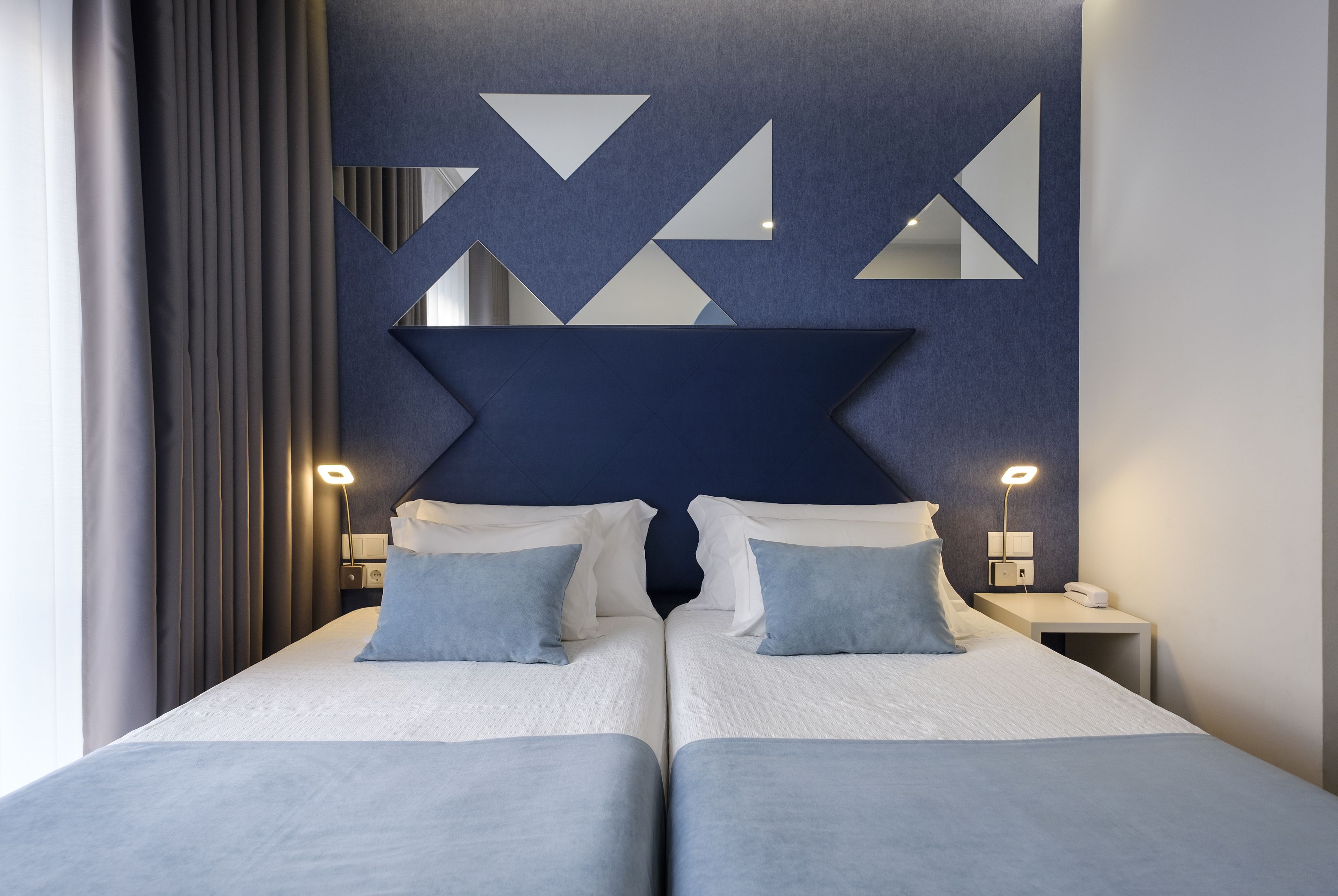 Uma cama de casal com duas almofadas, uma mesa de cabeceira com abajur e um design geométrico na parede.
