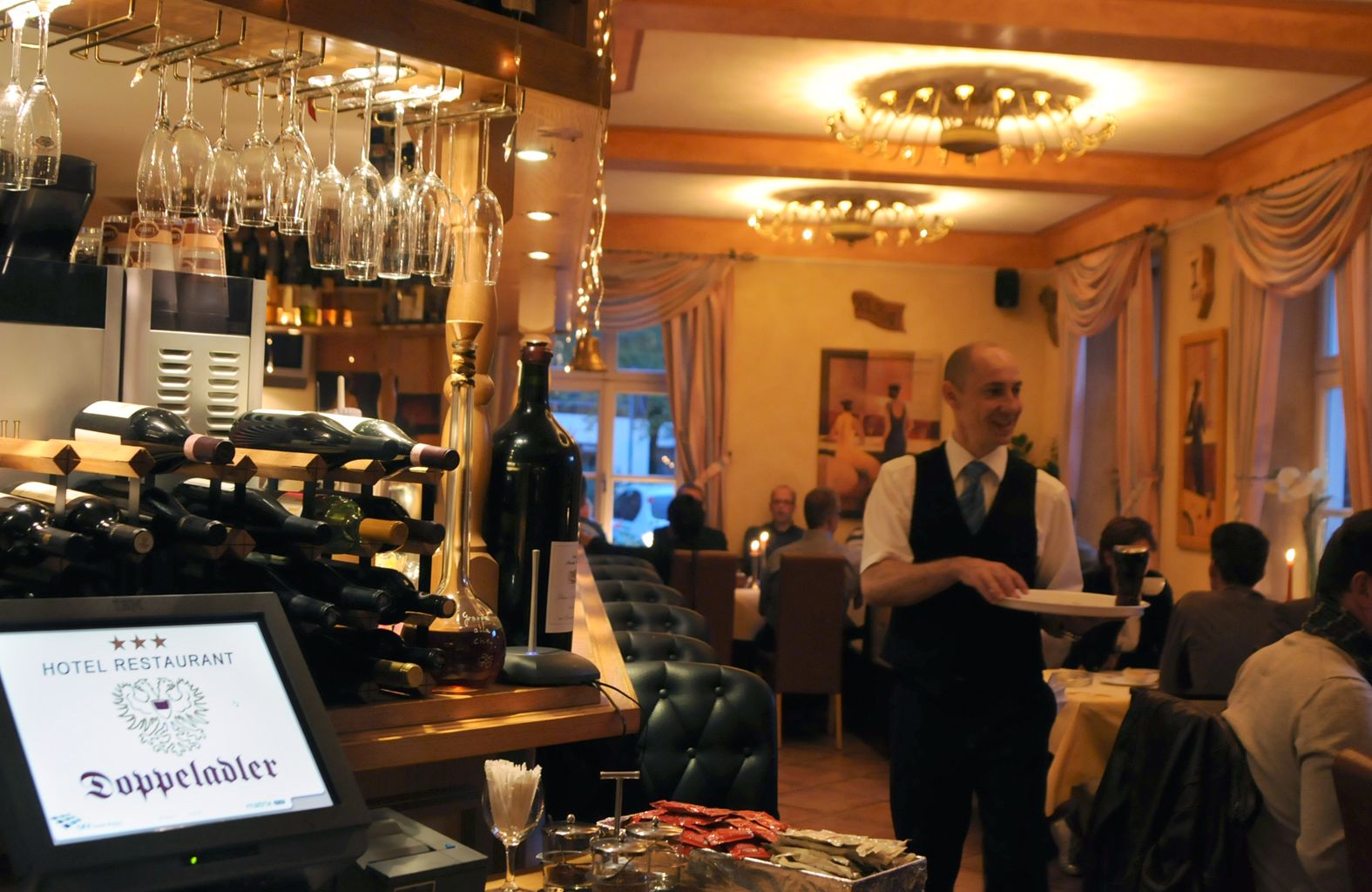 Interior de restaurante de hotel com área de bar, garrafas de vinho e um garçom atendendo os hóspedes.